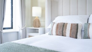 Cama bem arrumada com lençóis brancos e almofadas listradas, luminária de cabeceira e janela com cortinas.