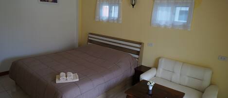 Quarto com cama, sofá, mesinha e duas janelas com cortinas.
