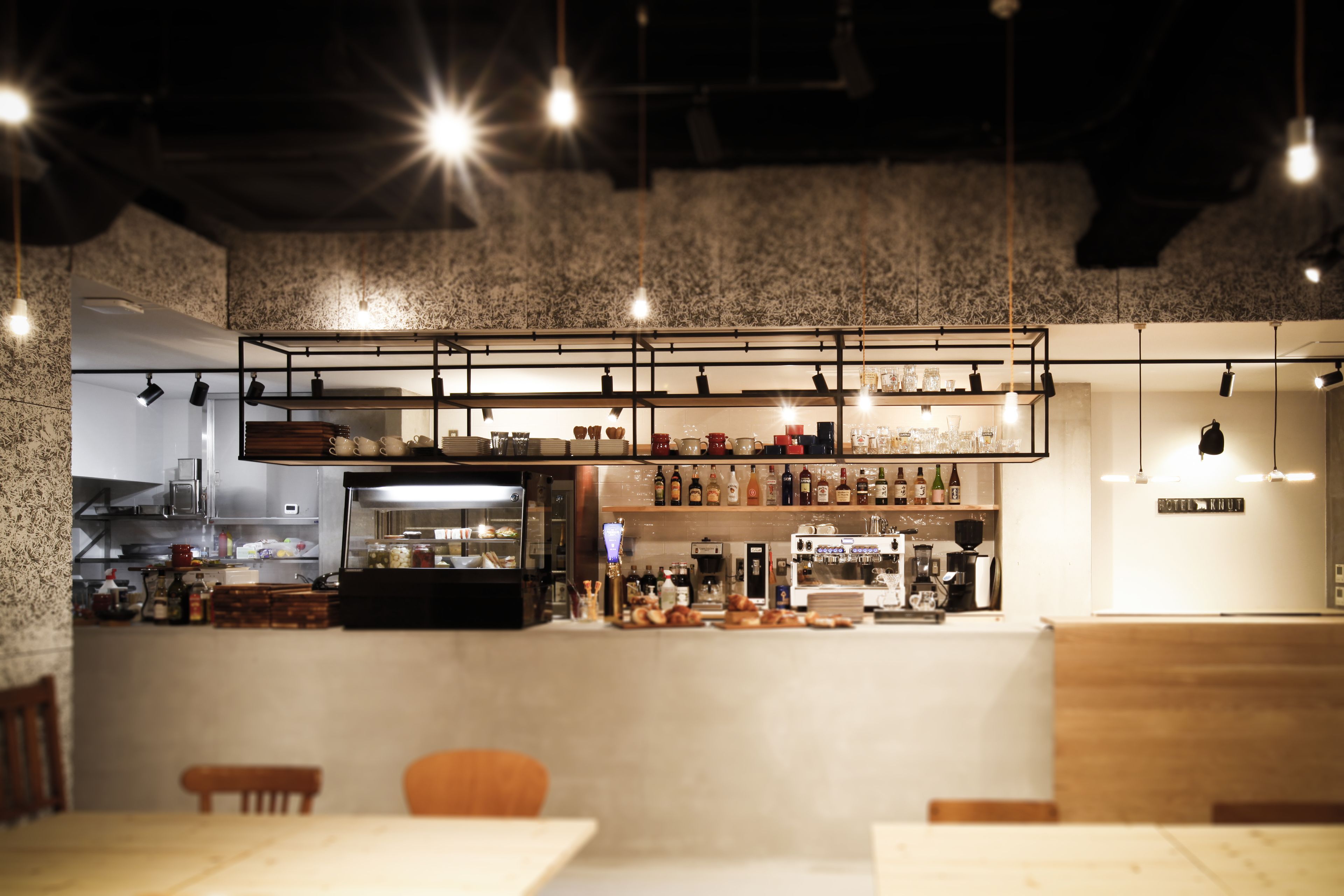Um restaurante com área de bar, mesas e cadeiras, e cozinha ao fundo.
