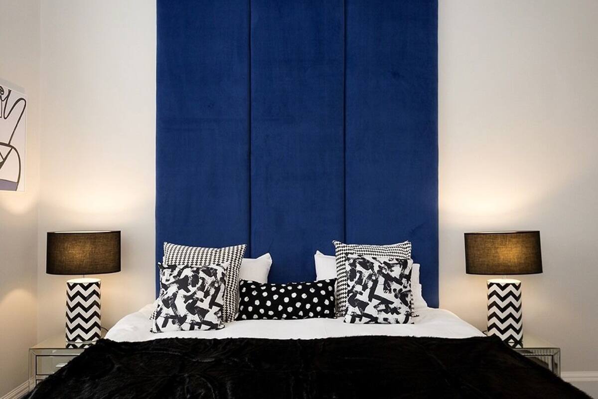 Quarto com cabeceira azul grande, edredom preto e almofadas estampadas. Há duas mesas de cabeceira com abajures.