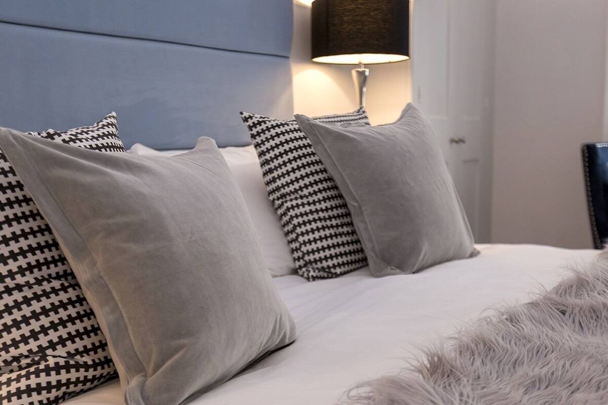 Uma cama bem arrumada com almofadas estampadas em cinza, preto e branco, um cobertor cinza fofinho e um abajur.