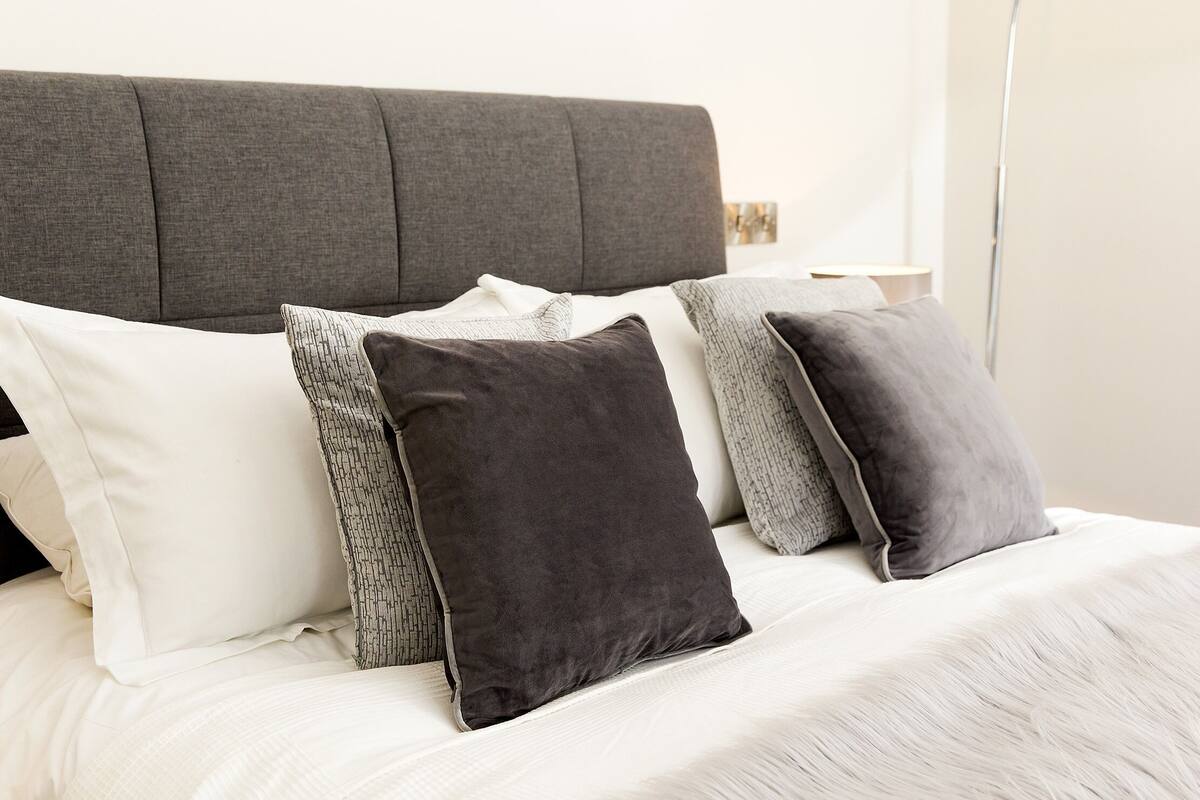 Cama com cabeceira estofada cinza, roupa de cama branca e vários travesseiros em diferentes texturas e cores.