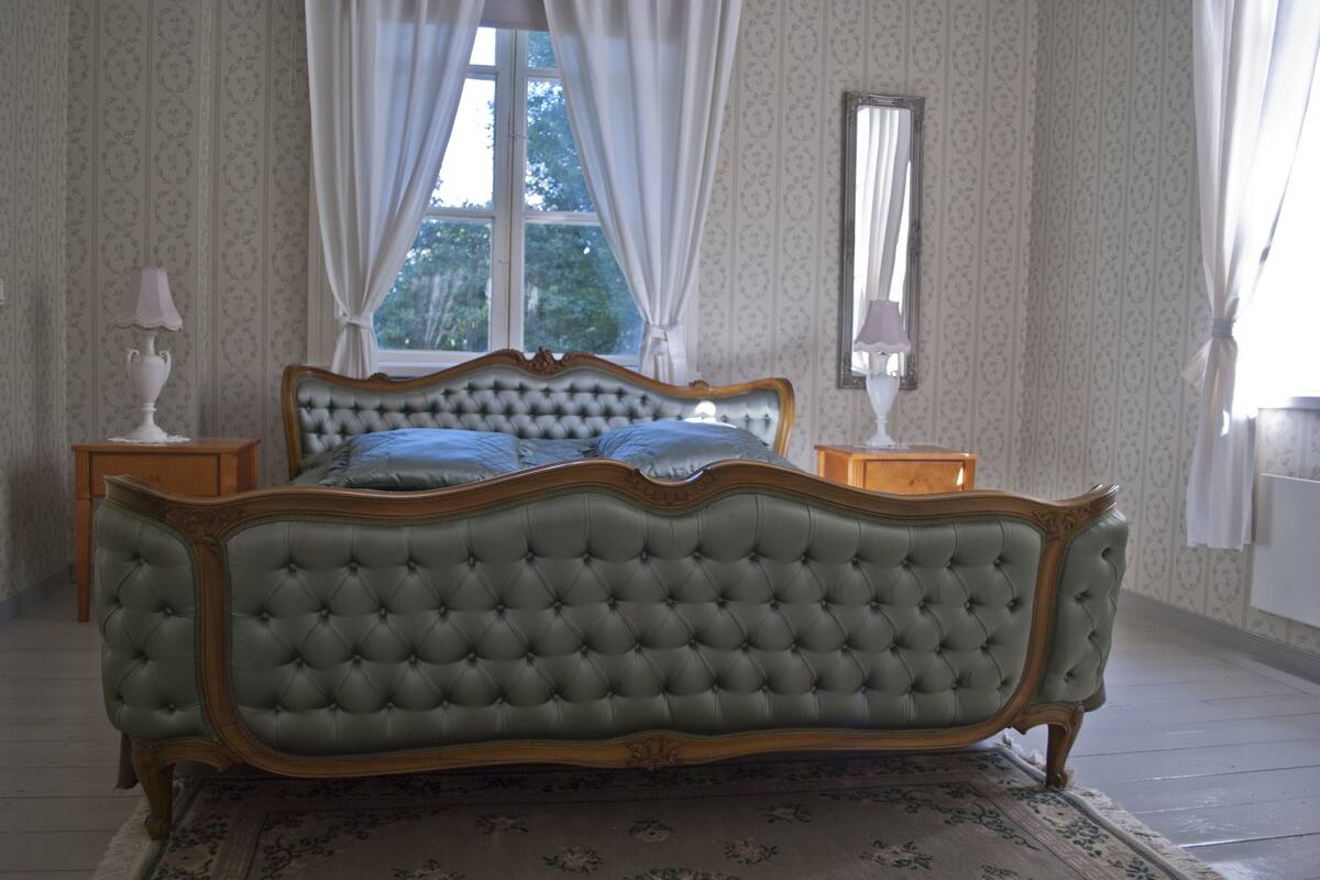 Quarto com cabeceira estofada, uma janela grande com cortinas, um espelho e duas mesas de cabeceira com abajures.