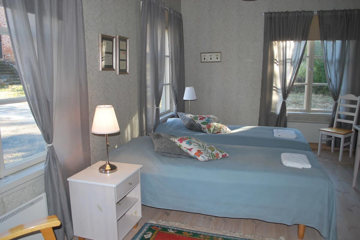 Quarto com duas camas, uma mesa de cabeceira com abajur, uma cadeira e uma janela com cortinas.