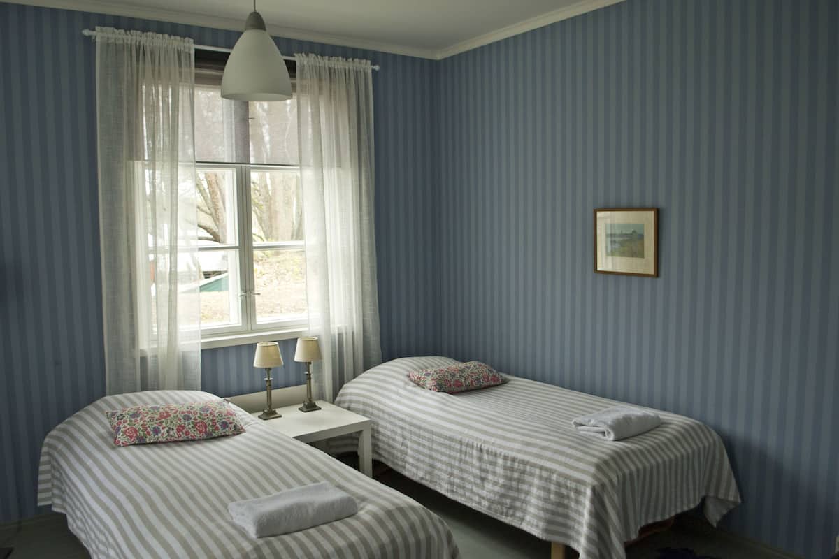 Quarto com duas camas, janela com cortinas, mesa de cabeceira com abajur e um quadro na parede.