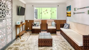 Sala de estar com móveis de vime, televisão de tela plana, janela com adesivos de borboletas e uma pintura de pôr do sol.