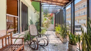 Uma área de estar coberta ao ar livre com um banco de madeira e uma cadeira de balanço, rodeada por plantas em vasos e um chão de azulejos de pedra.