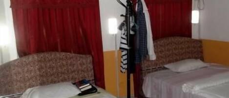 Quarto com duas camas, uma com cabeceira estampada e outra com cabeceira lisa. Há um guarda-roupa com roupas penduradas e uma pequena mesa com um livro e um abajur.