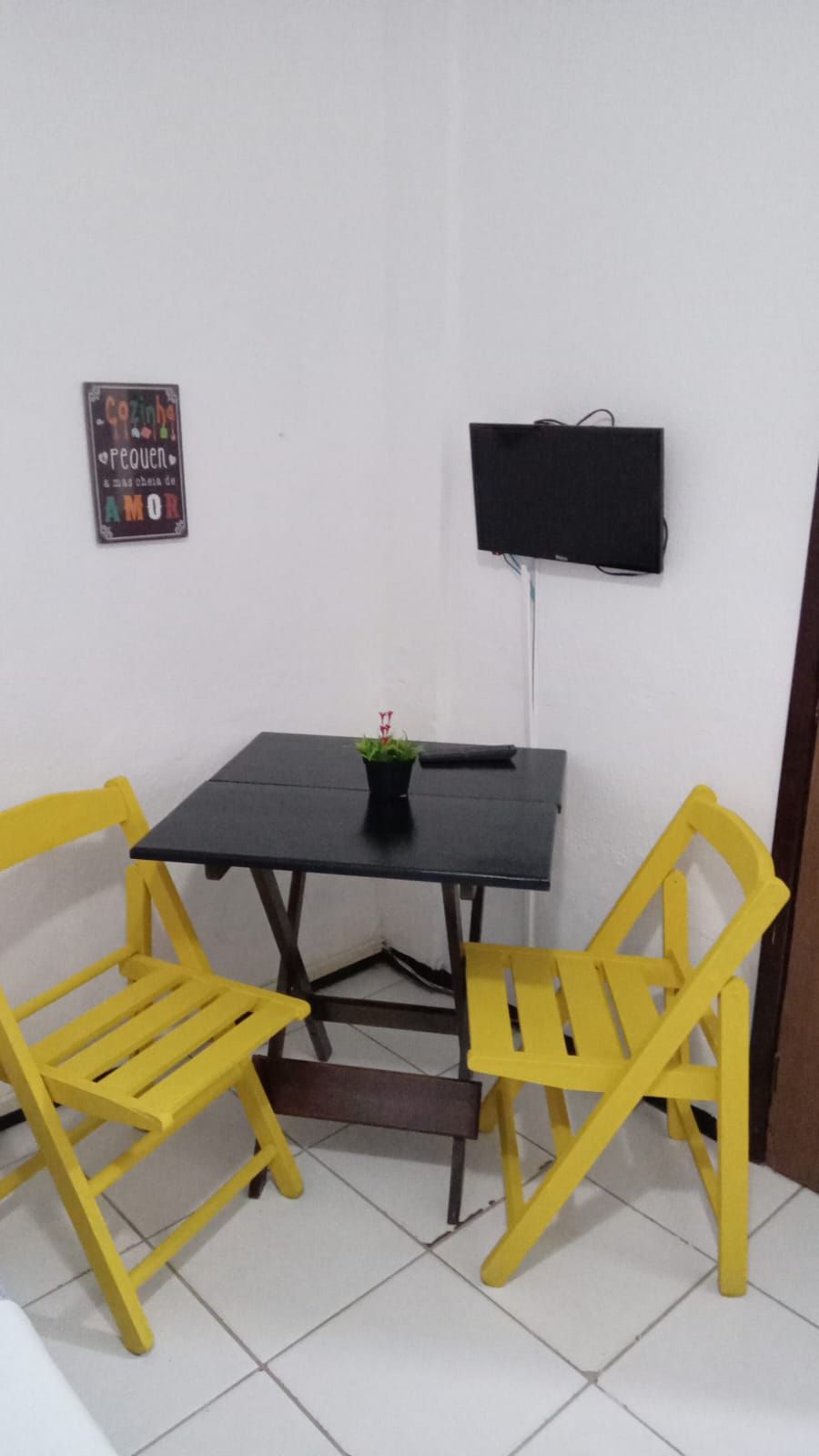 Uma pequena área de refeições com uma mesa preta, duas cadeiras amarelas, um vaso de plantas, uma TV na parede e um cartaz.