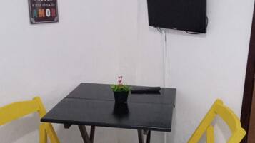 Uma pequena área de refeições com uma mesa preta, duas cadeiras amarelas, um vaso de plantas, uma TV na parede e um cartaz.