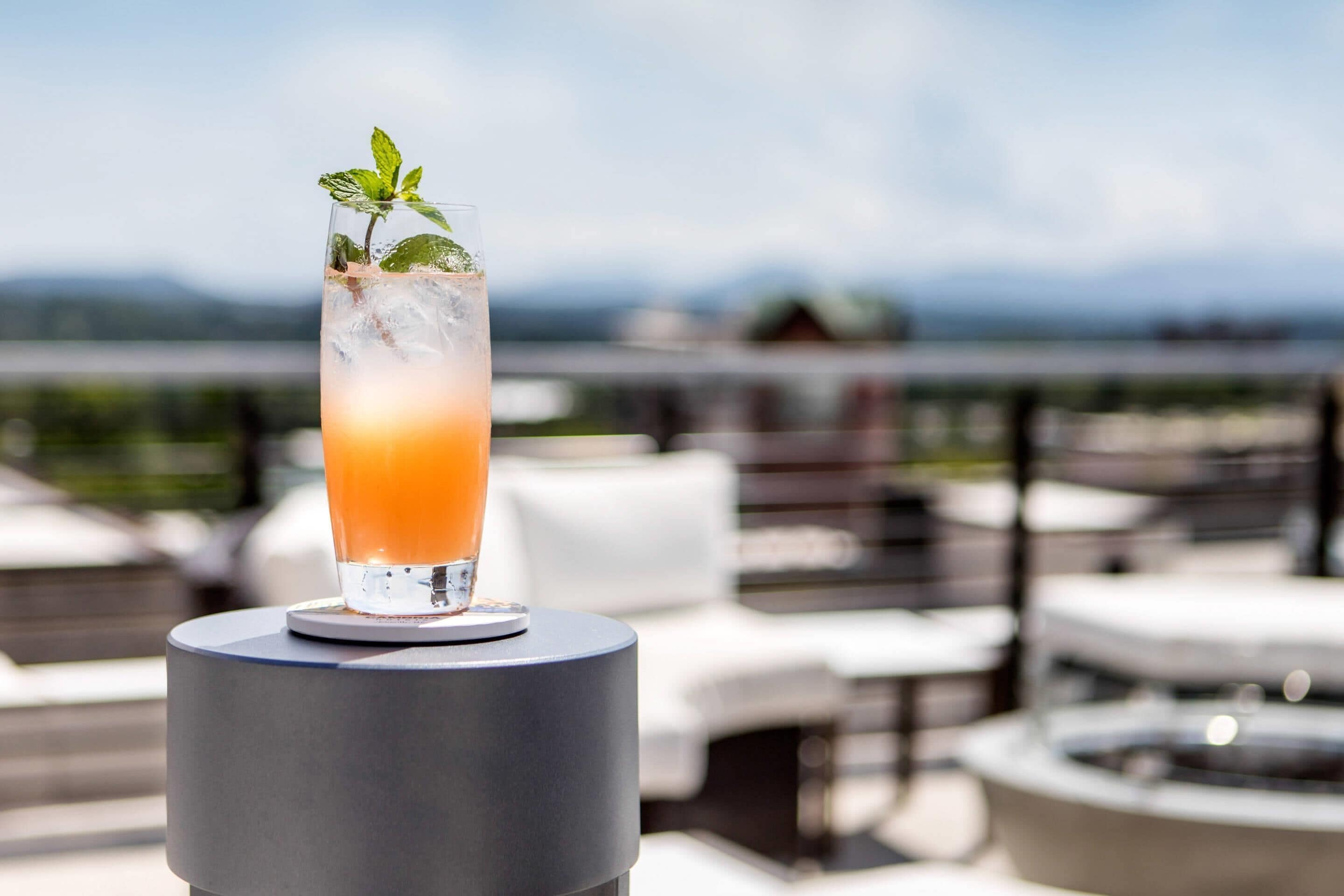 Um copo de bebida de laranja com enfeite de hortelã sobre uma mesa, em um terraço com vista para montanhas ao longe.