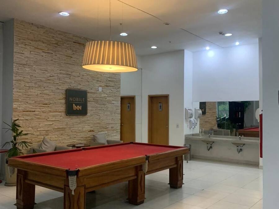 Uma mesa de bilhar em um saguão de hotel, com parede de pedra, uma placa com 'NOBLE INN' e uma luminária pendente moderna.