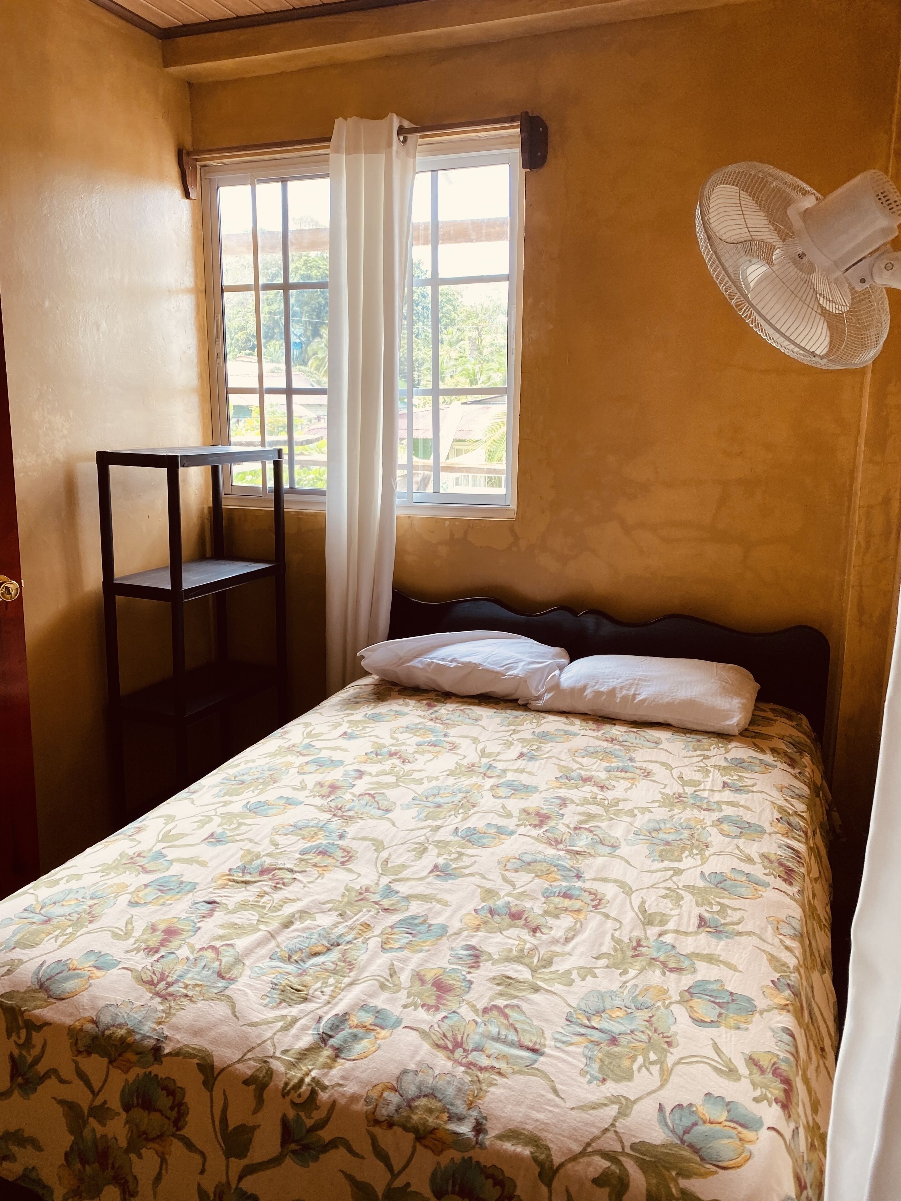 Quarto com uma cama, um ventilador, uma janela com cortinas e uma pequena mesa.