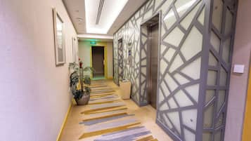 Corredor de hotel com carpete estampado, um vaso de planta, um extintor de incêndio e a porta de um elevador de vidro.
