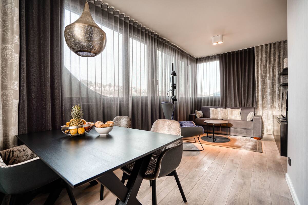 Sala moderna com mesa de jantar de madeira escura, um sofá e uma mesa de centro. Há uma janela grande com cortinas leves e vista para a cidade.