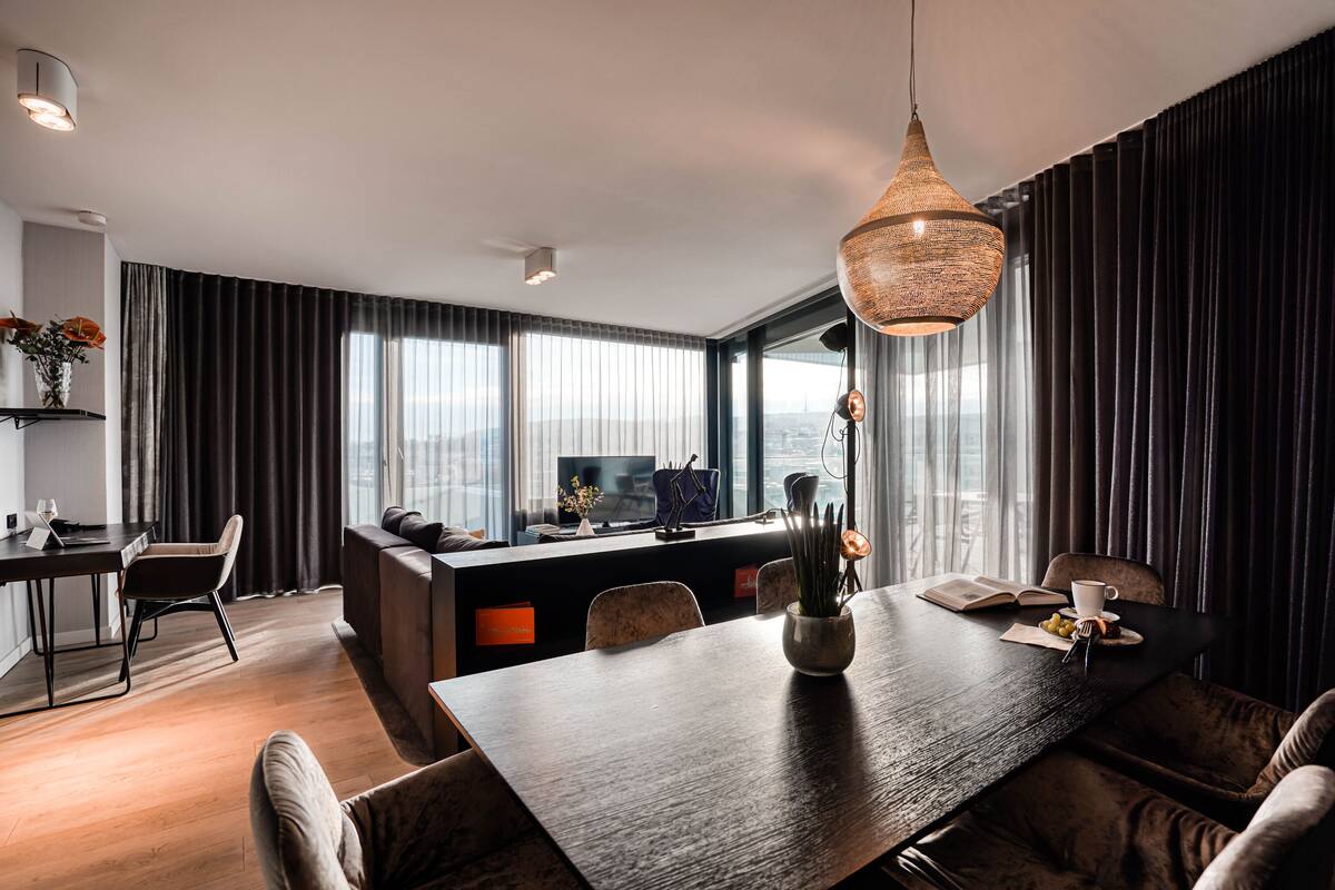 Quarto de hotel moderno com uma mesa de jantar em madeira escura, assentos confortáveis, uma janela grande com cortinas e vista para a cidade.