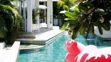 Piscina com boia rosa, rodeada por palmeiras e um edifício branco com colunas.