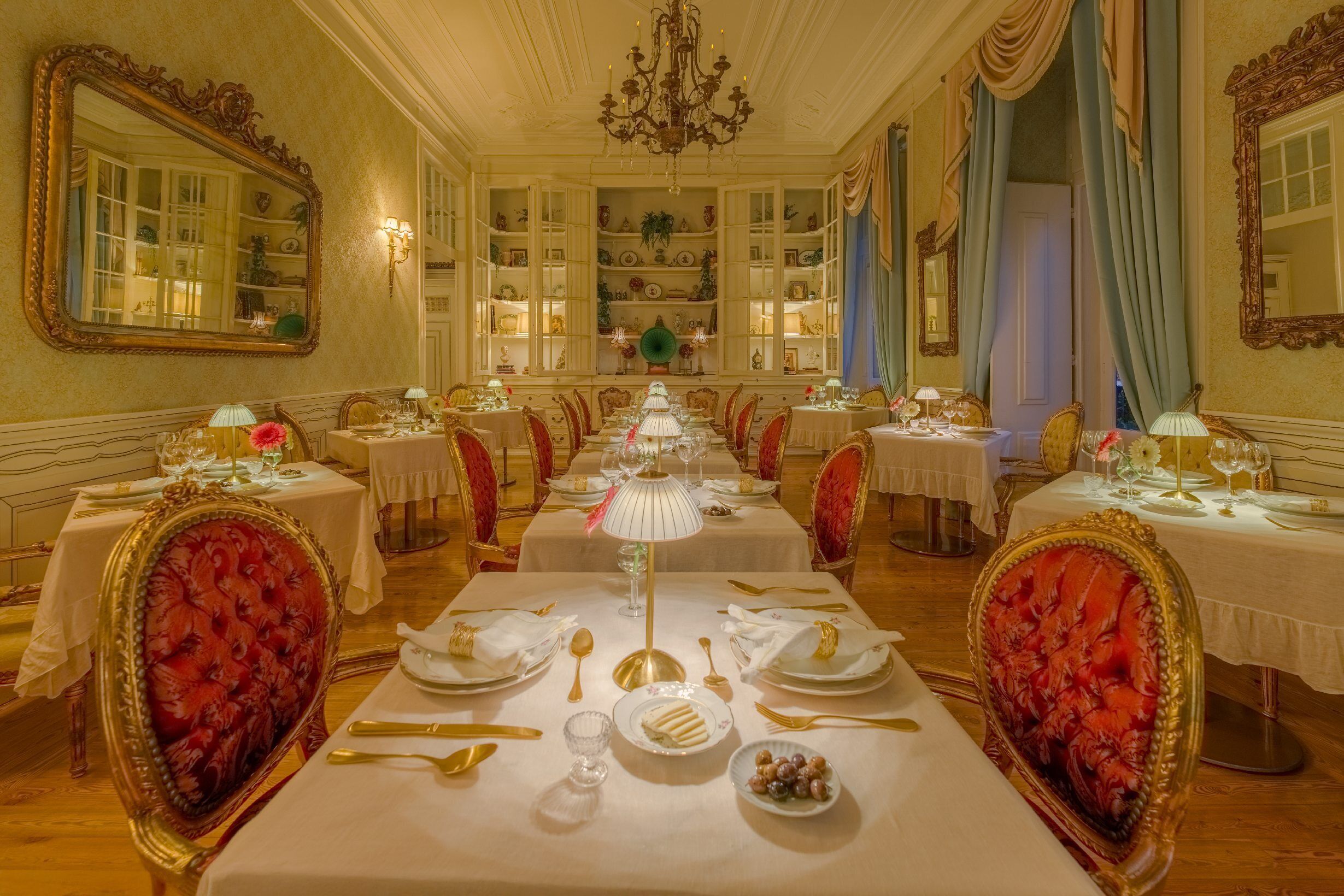 Uma sala de jantar formal com várias mesas postas para o serviço, cadeiras ornamentadas e um candelabro.