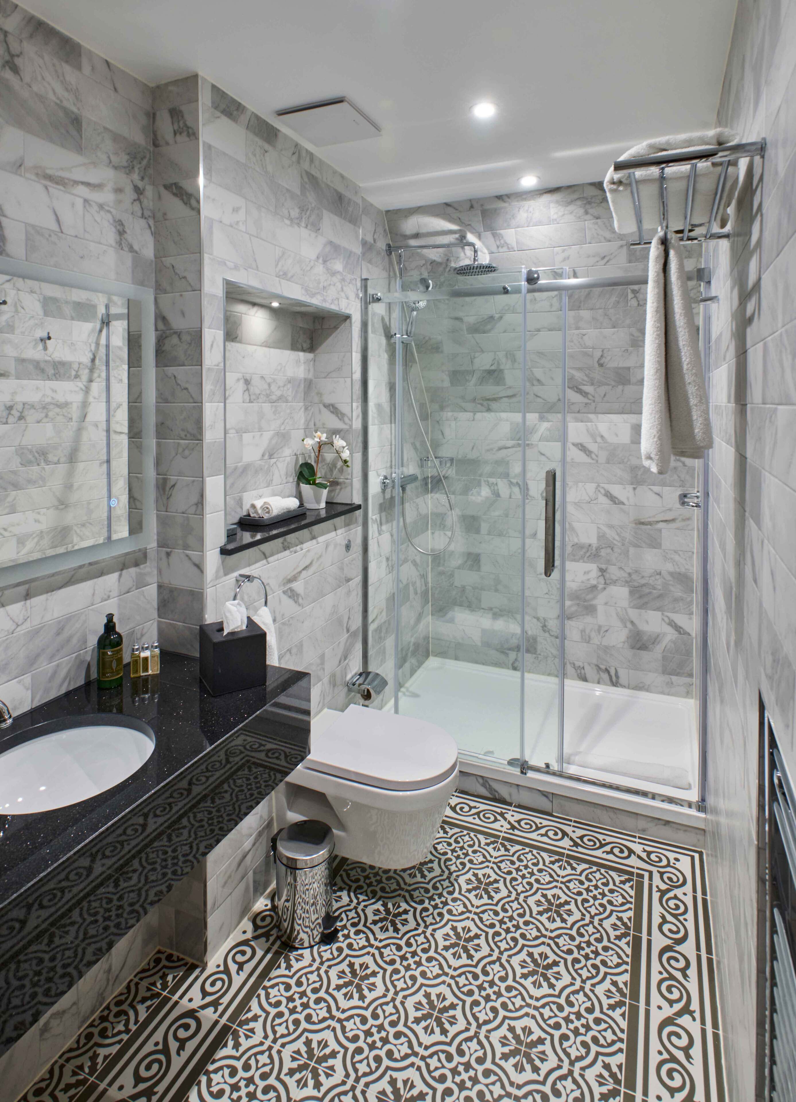 Um banheiro moderno com painel de backsplash de mármore, pia com bancada preta, vaso sanitário, chuveiro com box de vidro e piso com ladrilhos estampados.