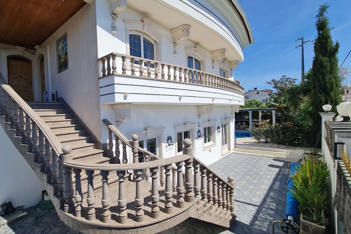 Um edifício de dois andares com uma varanda curva, uma escadaria com balaústres e um pátio pavimentado.