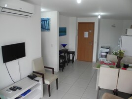 Quarto de hotel com televisão de tela plana, uma mesa com telefone, uma cadeira, área de refeições com mesa e cadeiras, cozinha com geladeira e porta com placa.