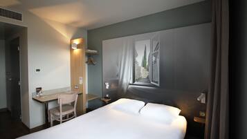 Quarto de hotel com cama, escrivaninha, cadeira, mesa de cabeceira e prateleira na parede.