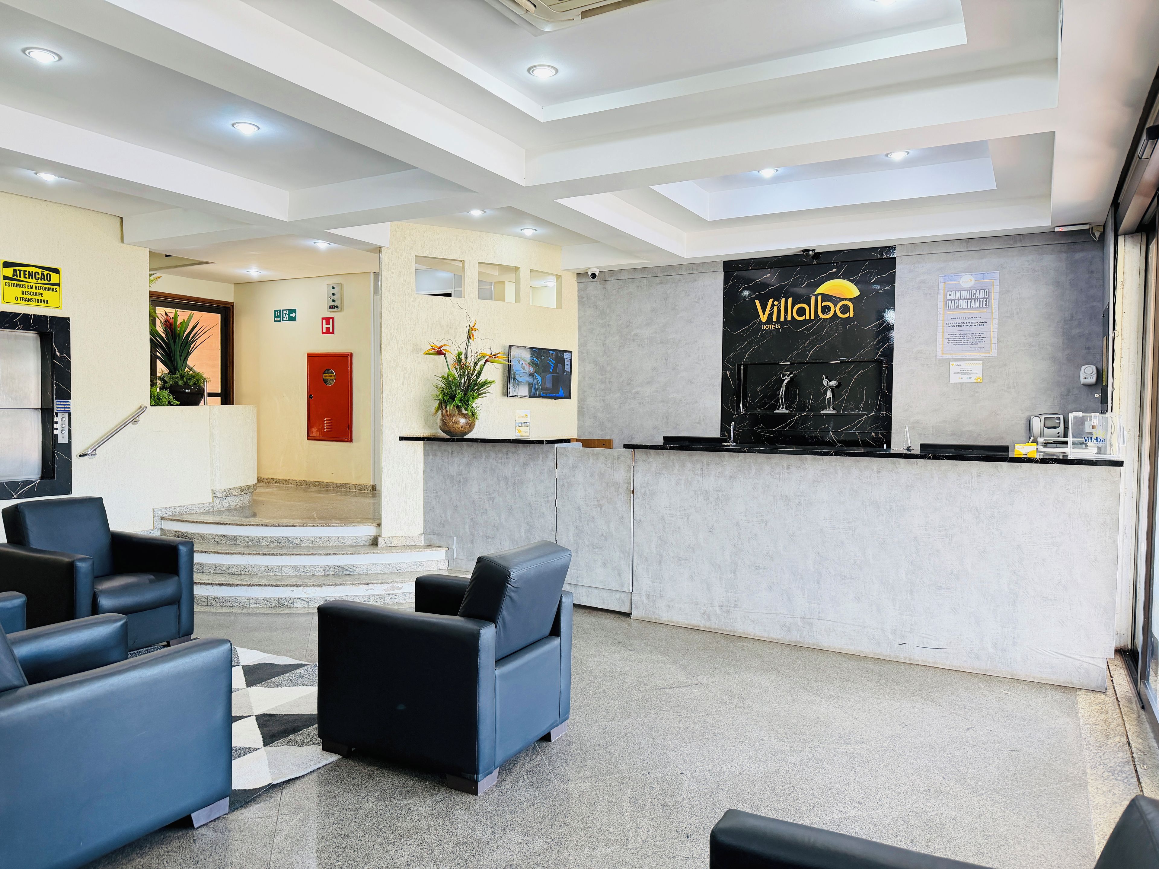 Uma área de recepção de hotel com balcão de mármore preto, mesa de atendimento e um espaço de espera com cadeiras azuis.