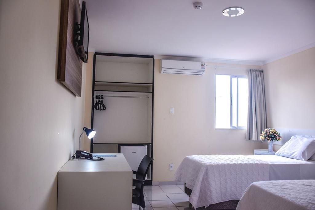 Quarto de hotel com escrivaninha, duas camas, guarda-roupa, ar condicionado e janela com cortinas.