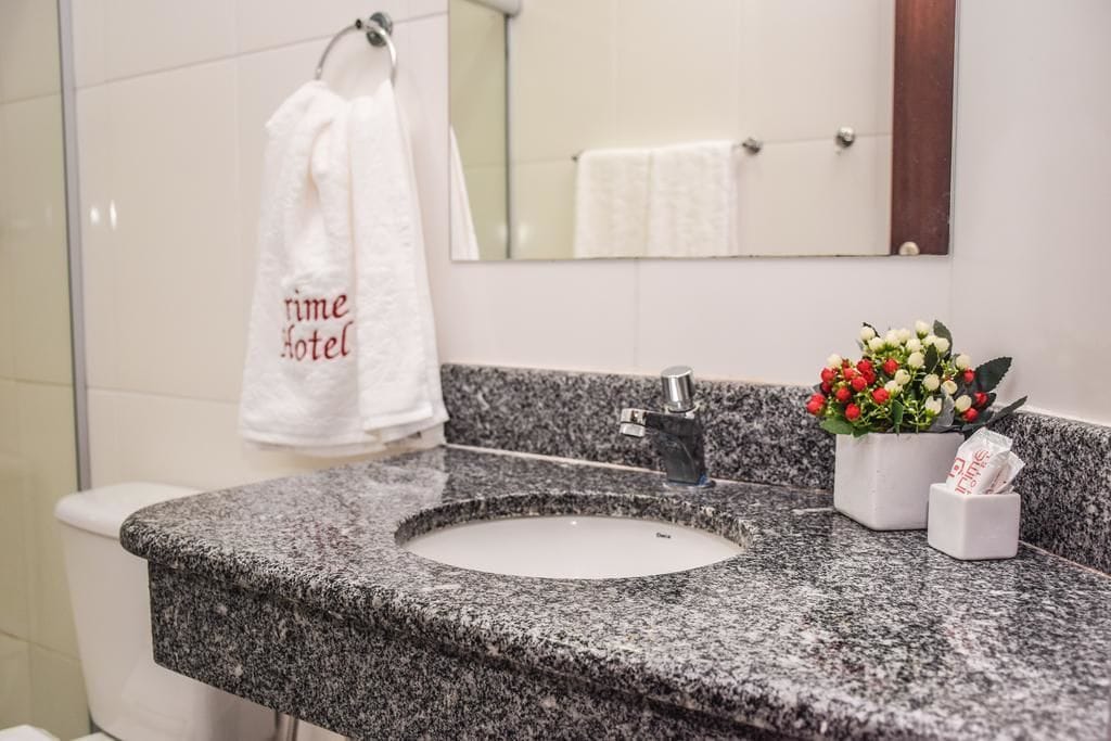 Casa de banho com lavatório de bancada em granito, espelho, porta-toalhas com toalha branca e uma pequena planta em vaso com flores vermelhas e brancas.