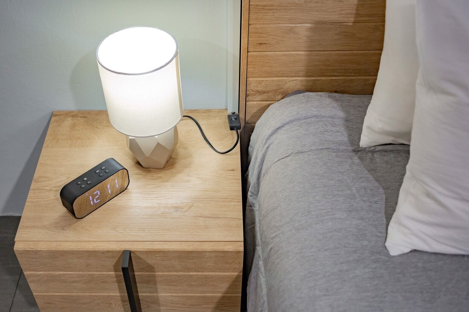 Uma mesa de cabeceira de madeira com uma lâmpada, um controle remoto e uma cama com almofadas.