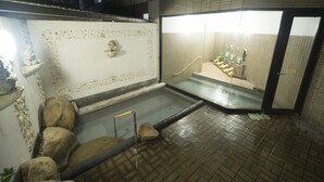 Uma onsen japonesa tradicional com banheira de pedra, uma pequena cascata e uma escadaria de madeira que leva a um vestiário.
