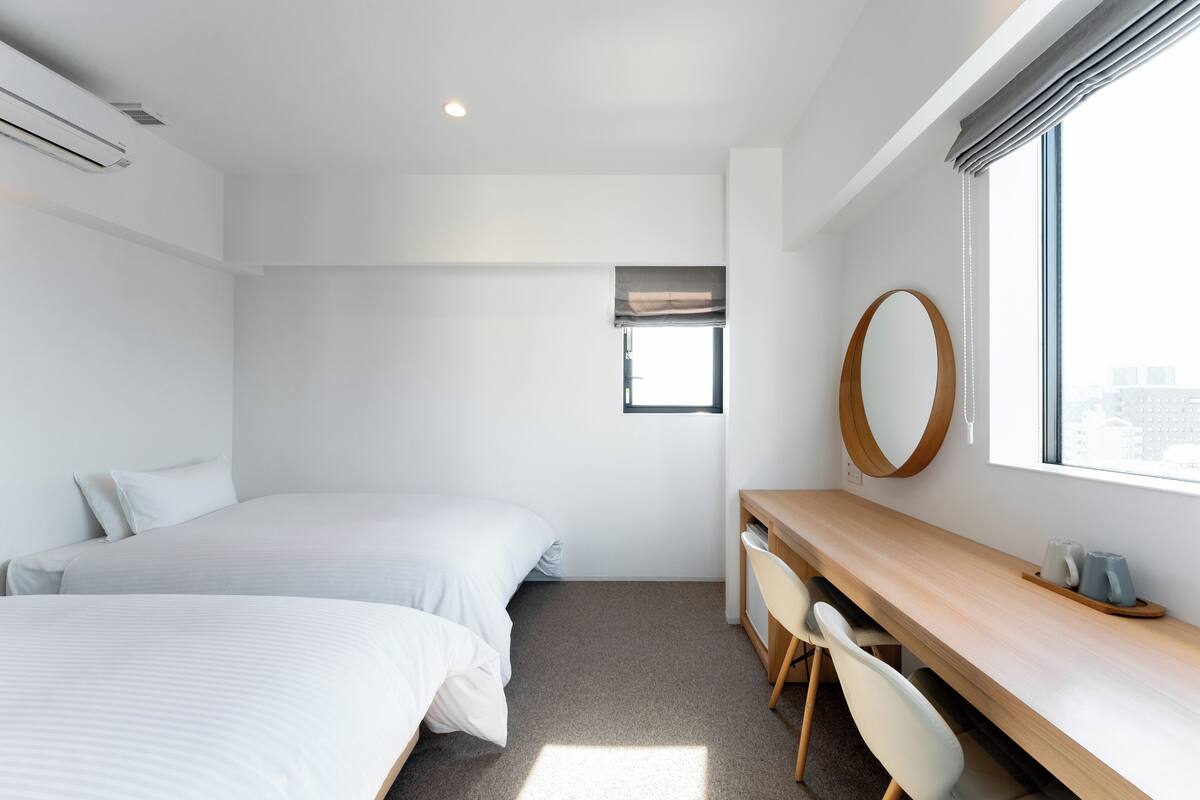 Quarto de hotel moderno com duas camas de solteiro, uma escrivaninha de madeira com cadeira, um espelho redondo e uma janela com vista para a cidade.