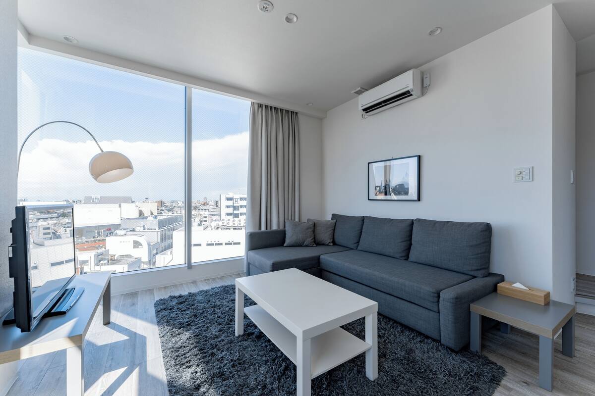 Uma sala de estar moderna com um sofá cinzento, uma mesa de centro branca e uma janela ampla com vista para a cidade.