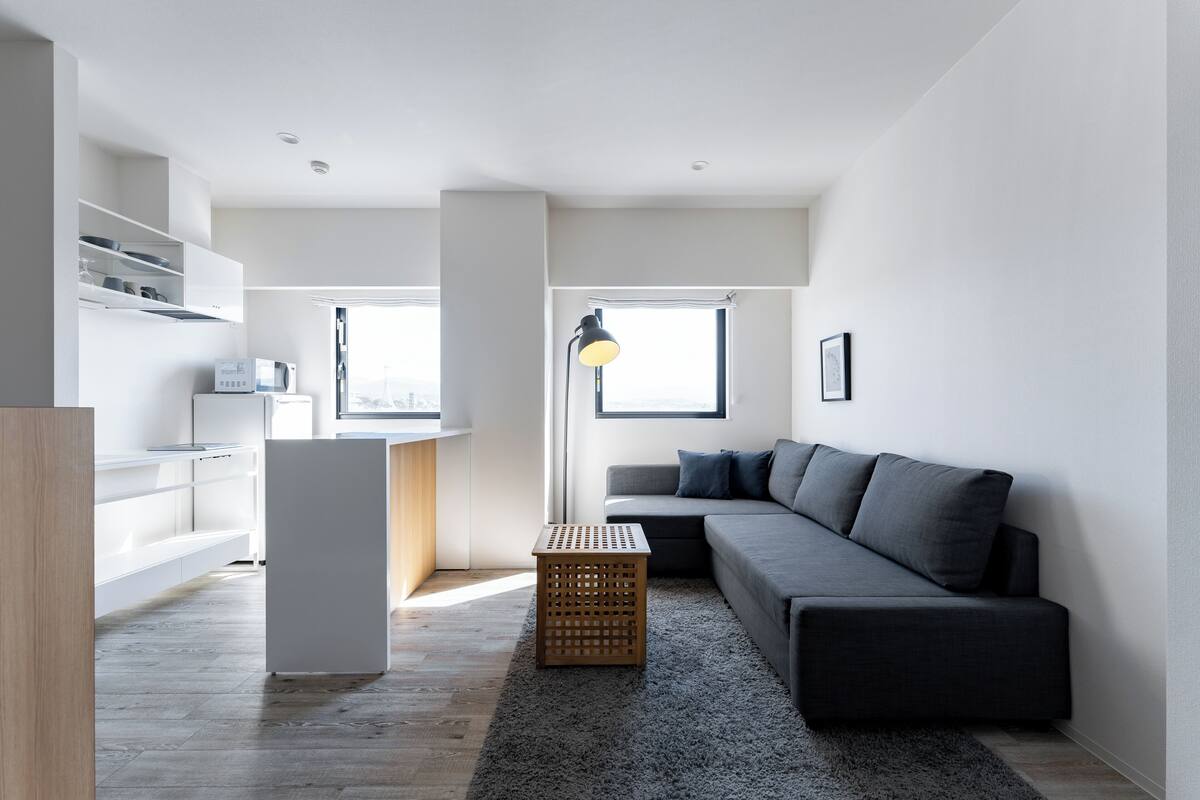Um espaço de estar moderno com sofá cinzento, uma pequena mesa de madeira e uma cozinha com armários brancos e micro-ondas.