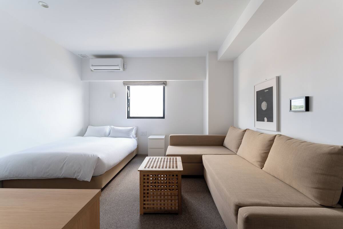 Quarto de hotel moderno com uma cama, um sofá, uma mesa de cabeceira de madeira e uma janela com persianas.