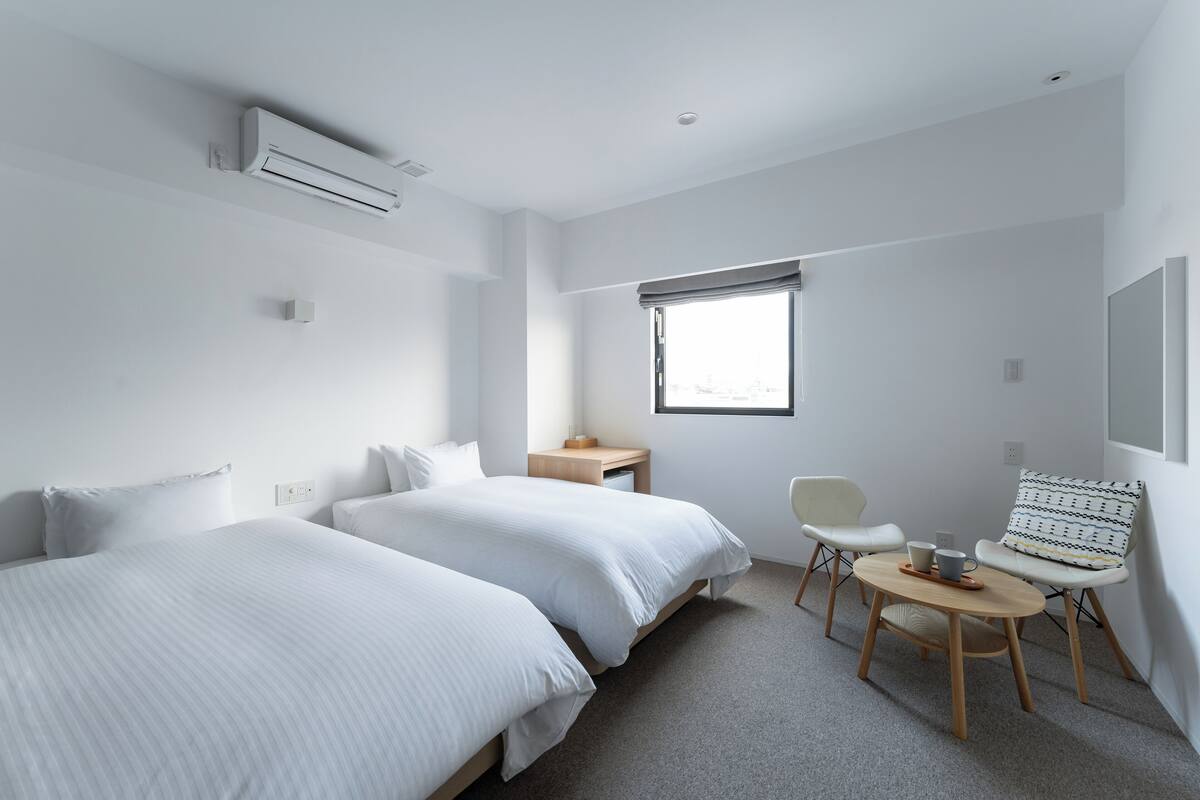 Quarto de hotel moderno com duas camas de solteiro, uma mesa pequena com uma cadeira, uma janela com vista e um ar condicionado.