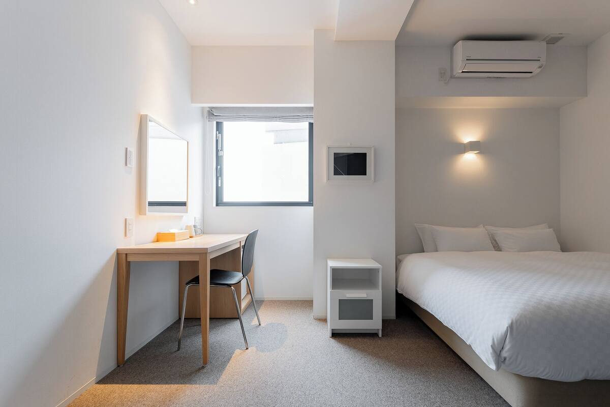 Quarto de hotel minimalista com uma cama, uma escrivaninha, uma cadeira, uma mesinha de cabeceira, ar condicionado na parede e uma janela com vista.