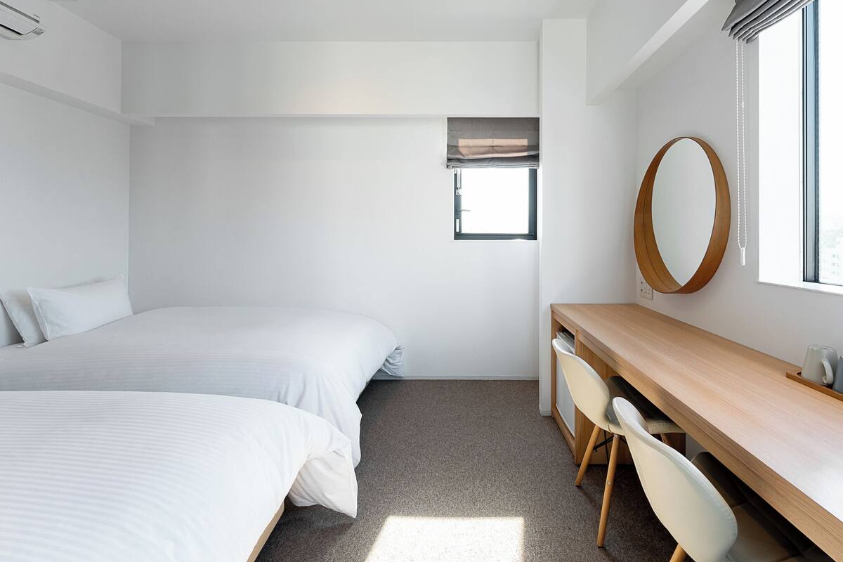 Quarto de hotel com duas camas, uma escrivaninha, uma cadeira, um espelho e uma janela.
