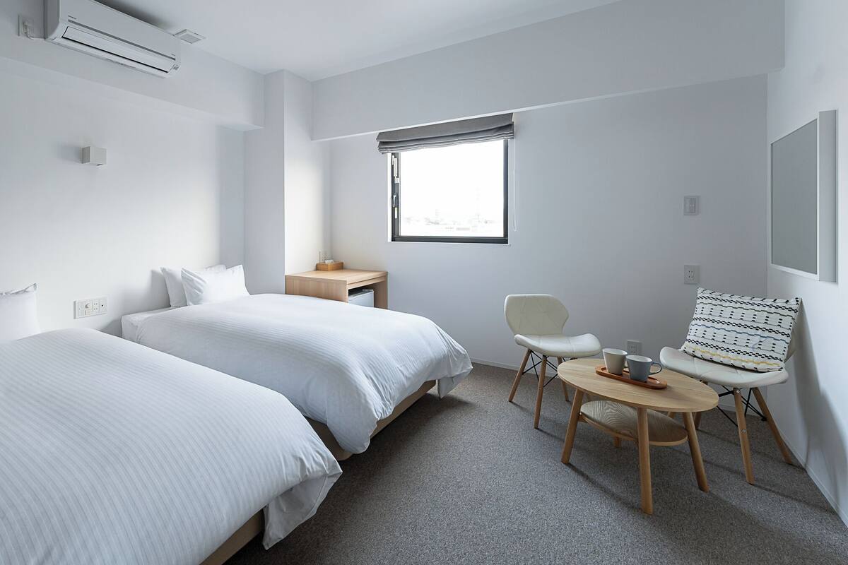 Quarto de hotel com duas camas, uma mesa pequena, uma cadeira, janela com vista e ar condicionado.