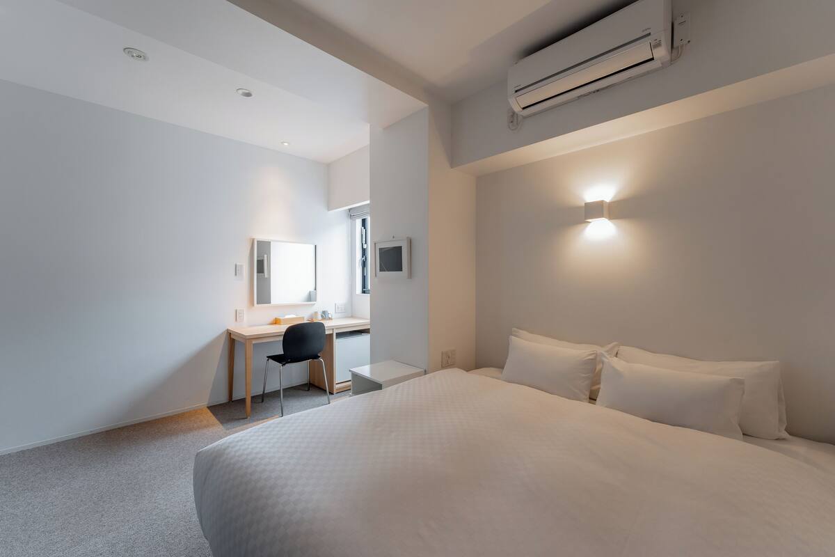 Quarto de hotel com uma cama grande, uma escrivaninha com cadeira, uma mesinha pequena e ar condicionado fixado na parede.