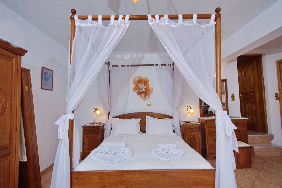 Uma cama com dossel e cortinas brancas, duas mesas de cabeceira com abajures e uma cabeceira de madeira.