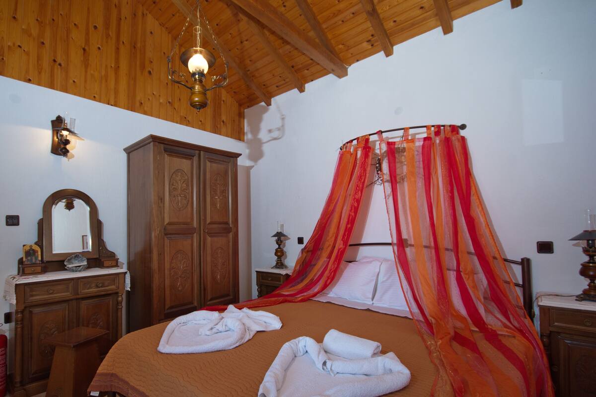 Quarto com teto de madeira, cama com dossel vermelho, guarda-roupa de madeira, mesa de cabeceira e um espelho.