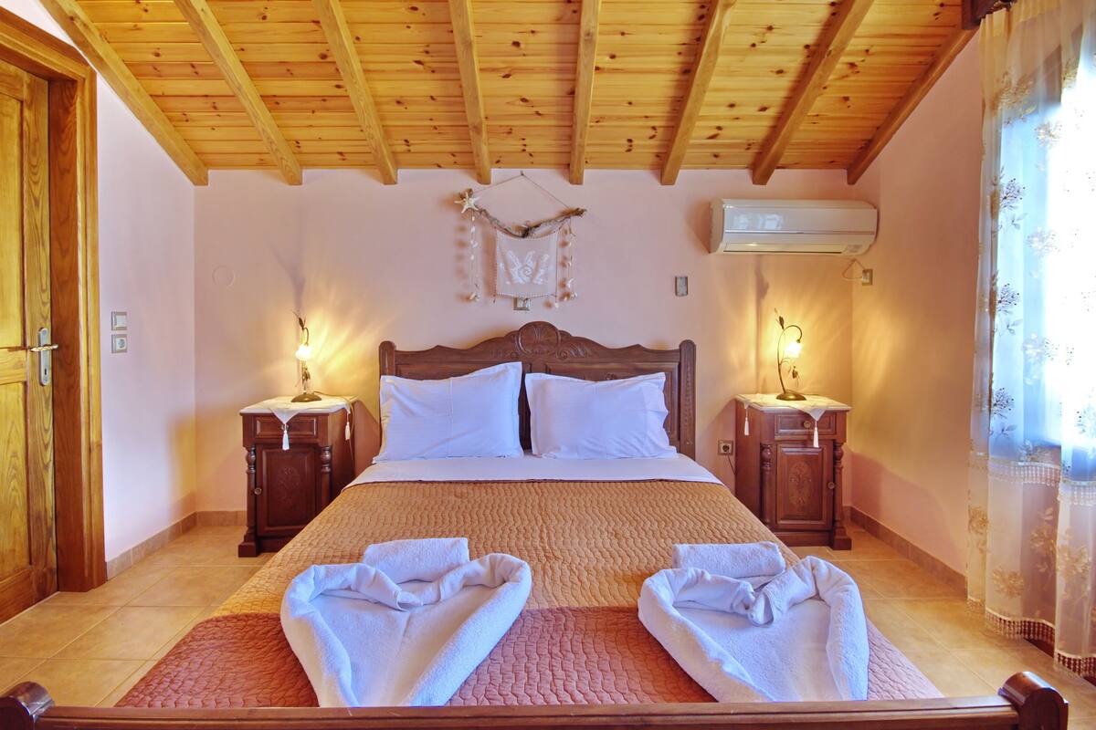 Quarto com teto de madeira, cama com roupa de cama branca, duas mesas de cabeceira com abajures e uma janela com cortinas florais.