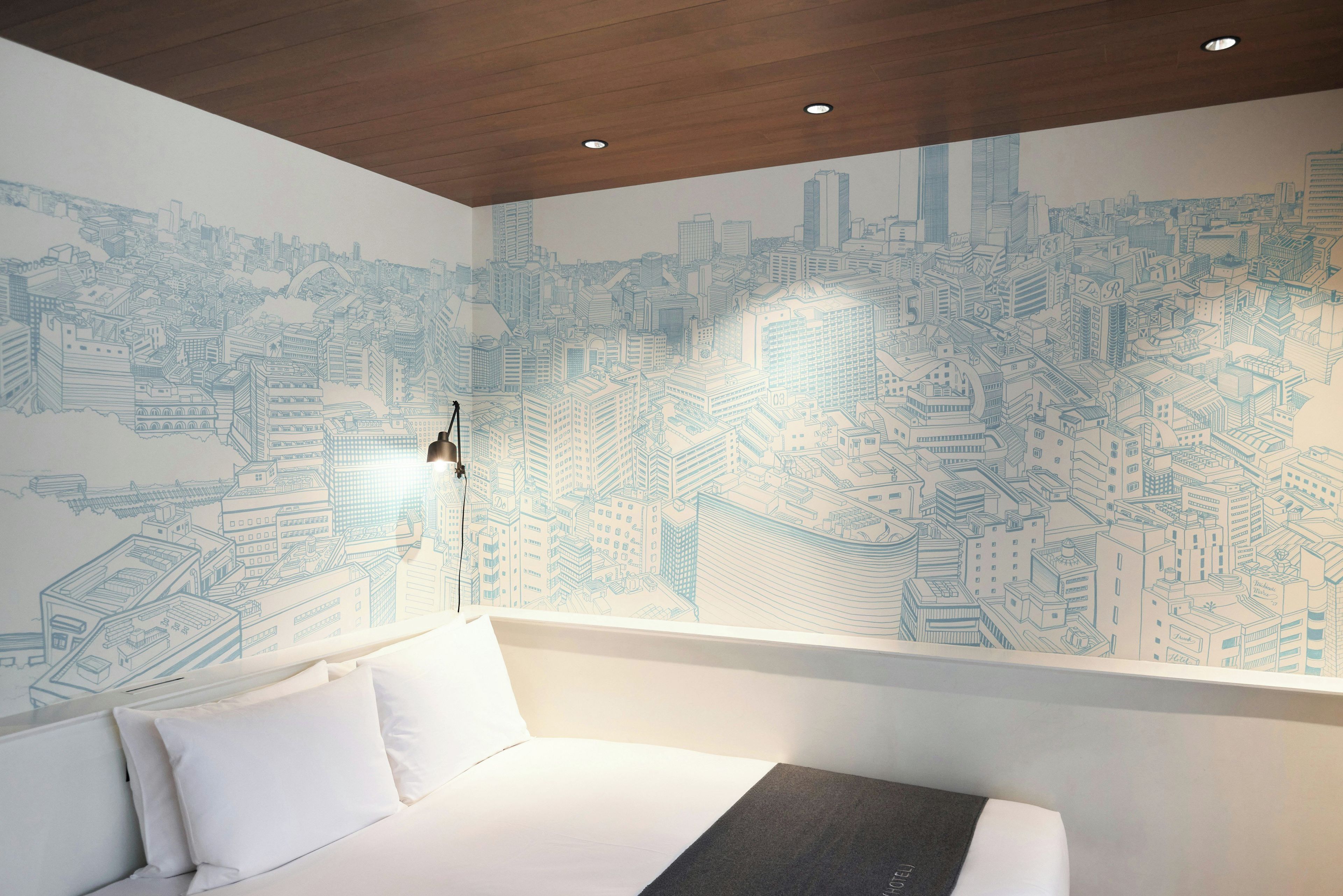 Quarto de hotel com um grande mural da paisagem urbana na parede, uma cama branca com almofadas e teto de madeira com iluminação embutida.
