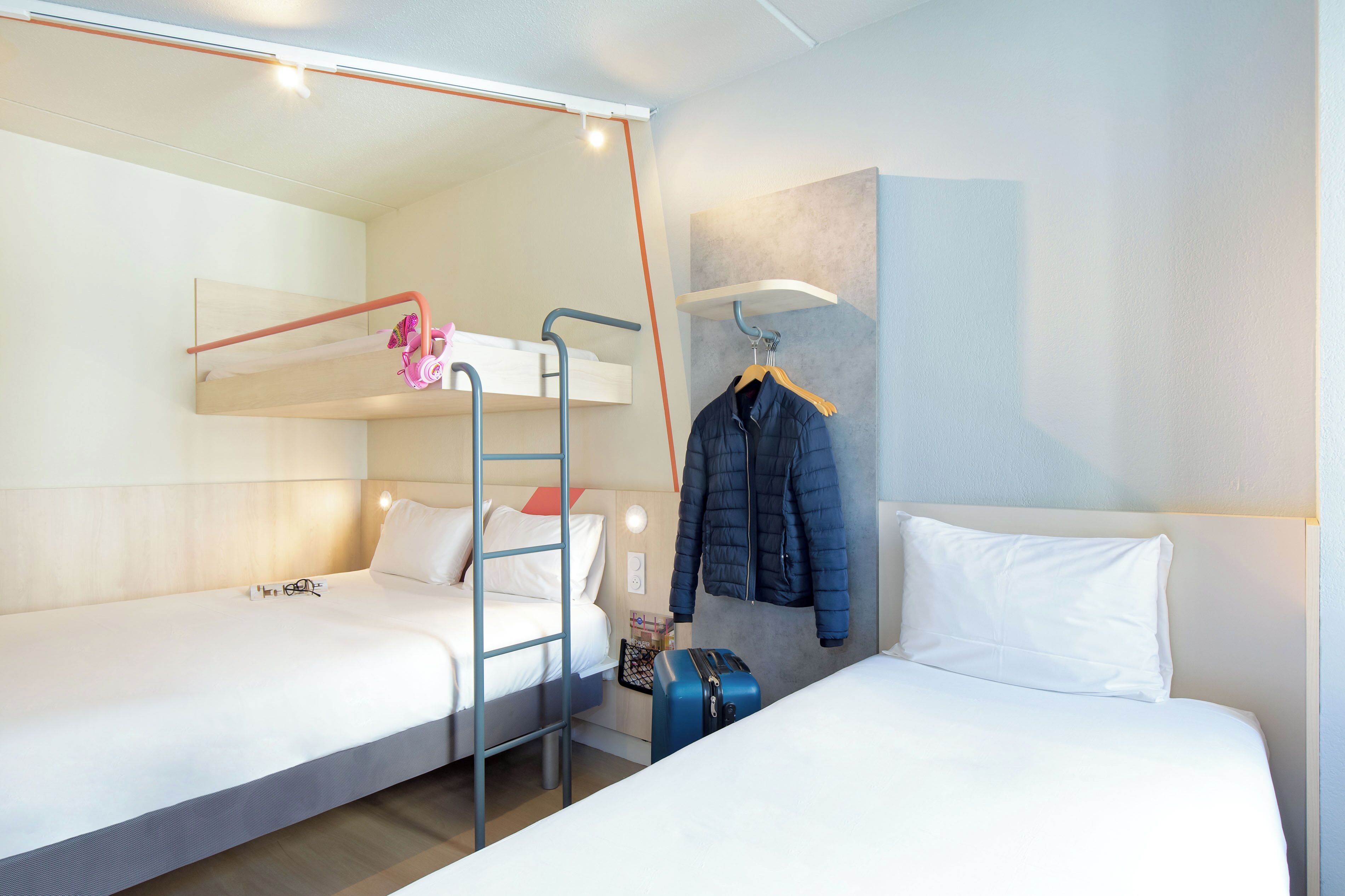 Quarto de hotel com beliches, cabideiro suspenso, mala e prateleira fixada na parede.