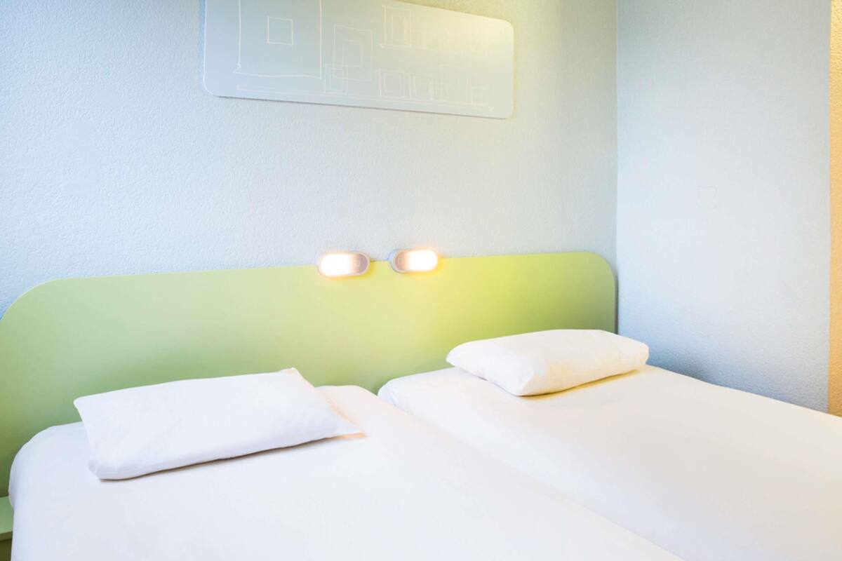 Quarto de hotel com duas camas de solteiro, cabeceira verde e duas luzes fixadas na parede.