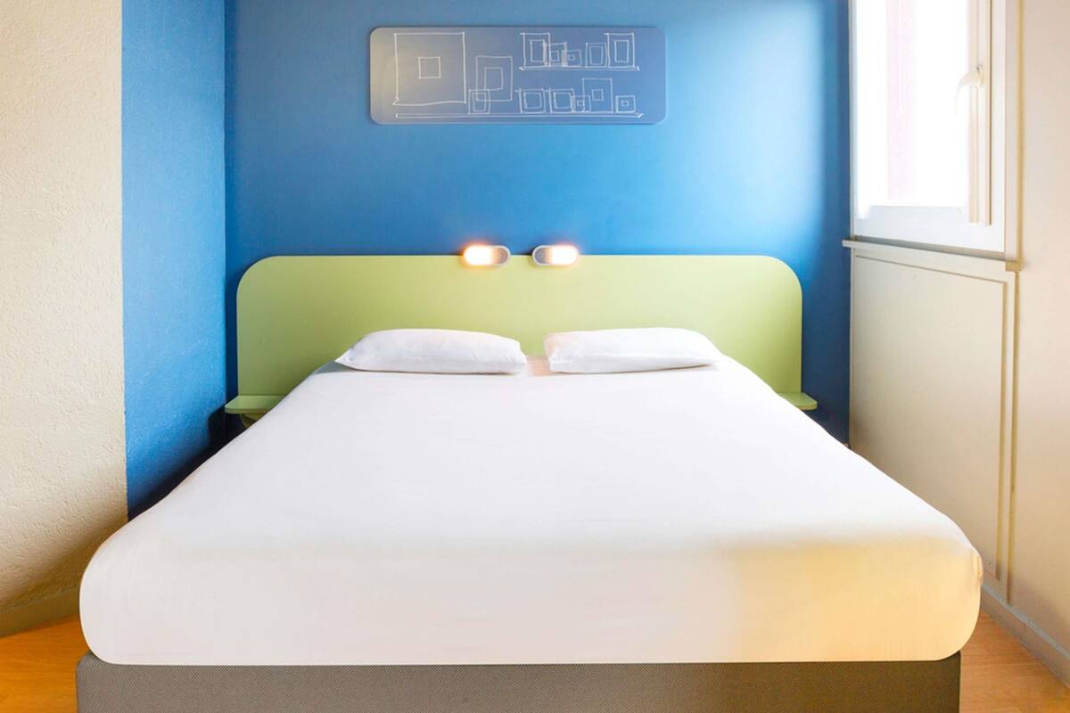 Uma cama com cabeceira verde, roupa de cama branca e duas luminárias de apoio.