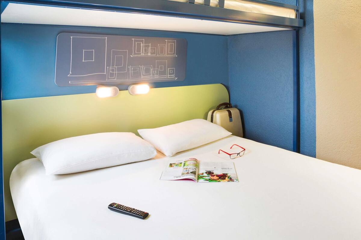 Quarto de hotel com cama beliche, mesa de cabeceira com controle remoto, uma revista e uma mala.