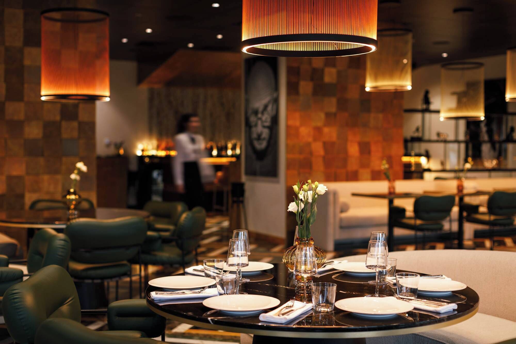 Ambiente de restaurante com mesas redondas preparadas para refeições, um garçom ao fundo e iluminação ambiente acolhedora.