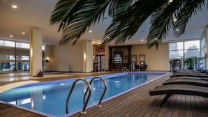 Piscina interior com deck de madeira, área de bar e vista da cidade através de grandes janelas.